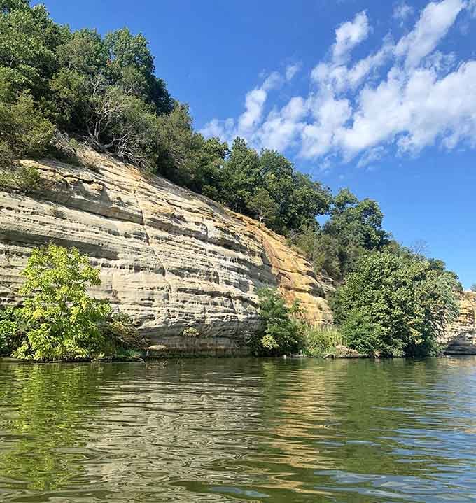 Striped limestone cliffs tower above the calm river waters, their geological layers visible like pages in Earth's diary.