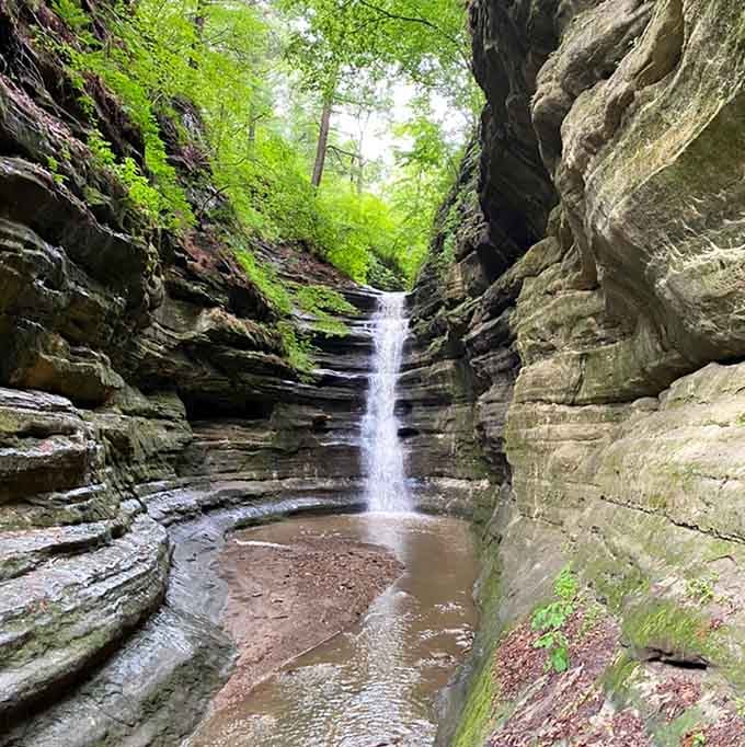 A hidden waterfall tucked between narrow canyon walls drops gently into a sandy pool surrounded by ancient stone.