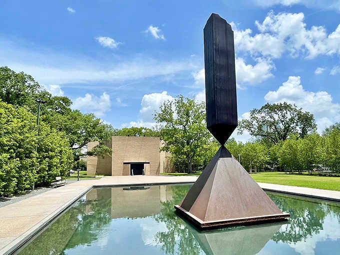 The stark sculpture rises from still water, creating a mirror image that doubles its contemplative power.
