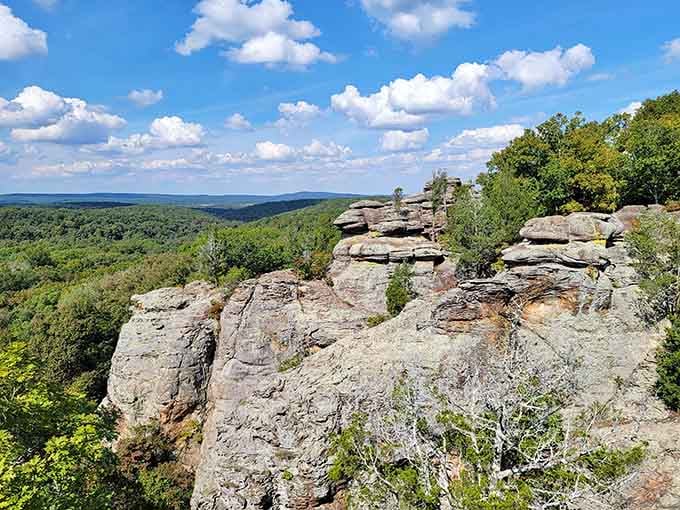 Layered rock formations create natural sculptures against blue skies and rolling green hills below.