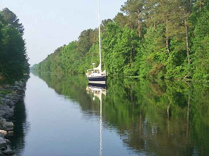 The sailboat rests peacefully in glass-smooth water reflecting the green canopy overhead in perfect symmetrical harmony.