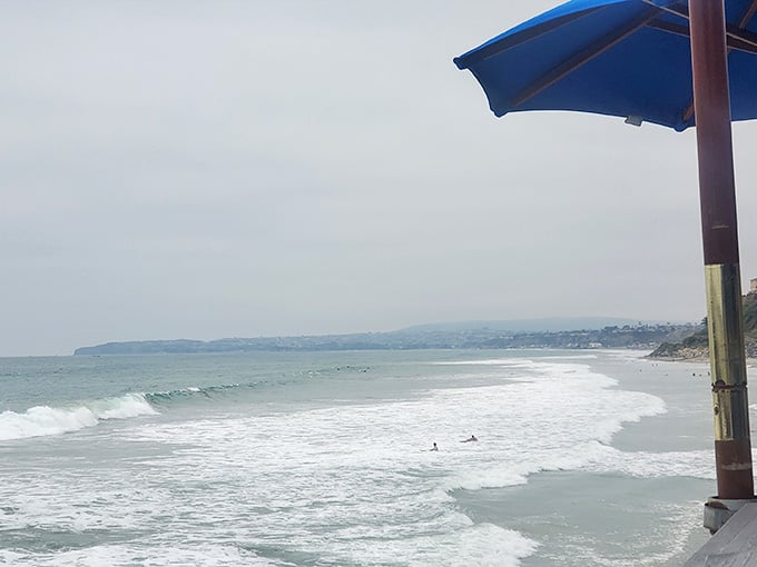 Under a blue umbrella, the Pacific stretches endlessly while surfers dot the waves like punctuation marks on water.