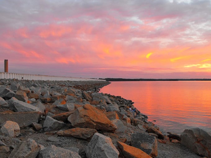 Sunset ignites the sky in brilliant coral and tangerine hues, turning the rocky dam into a front-row seat.