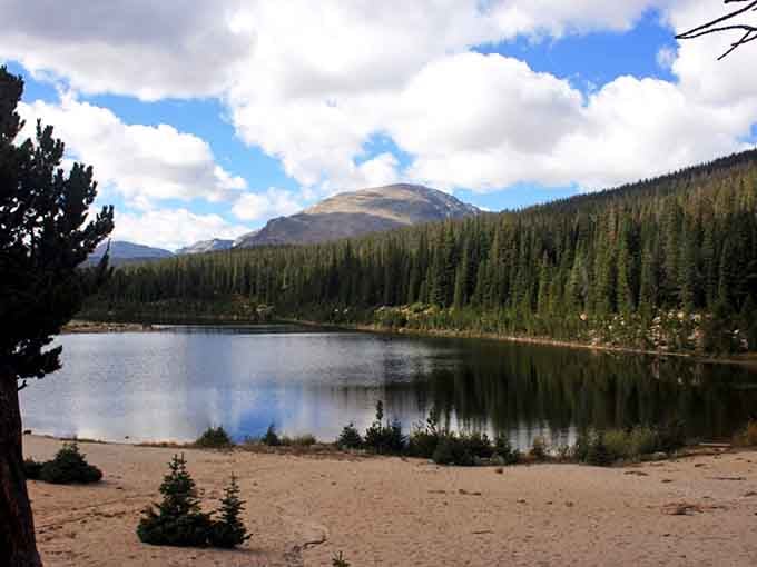 This stunning alpine beach sits beside crystal-clear water surrounded by pine forests and towering mountains after a rewarding hike.