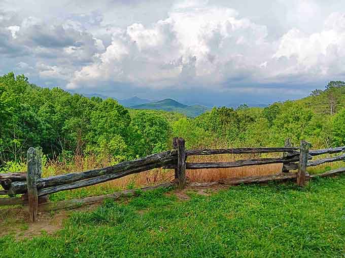 That old-fashioned fence frames vibrant spring greenery while towering clouds promise afternoon drama over the mountain wilderness.