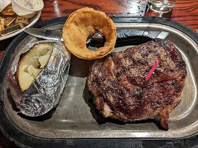 A perfectly cooked ribeye with baked potato and onion ring demonstrates why steak lovers make this place a regular pilgrimage destination.