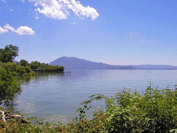 Clear mountain waters reflect perfect blue skies while gentle shoreline greenery frames this tranquil lakeside escape wonderfully.
