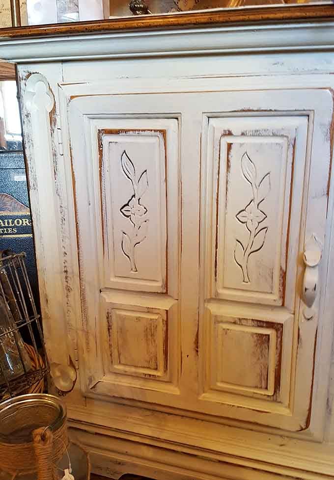 Hand-carved floral details on distressed white cabinetry showcase craftsmanship that modern furniture factories forgot how to replicate entirely.