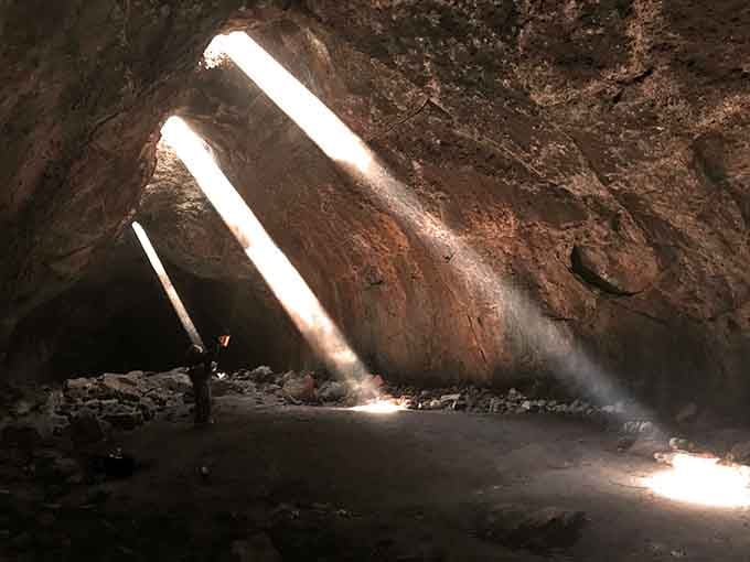Sunlight pierces the cave's ceiling in dramatic beams, illuminating the rocky floor like nature's own cathedral lighting design.