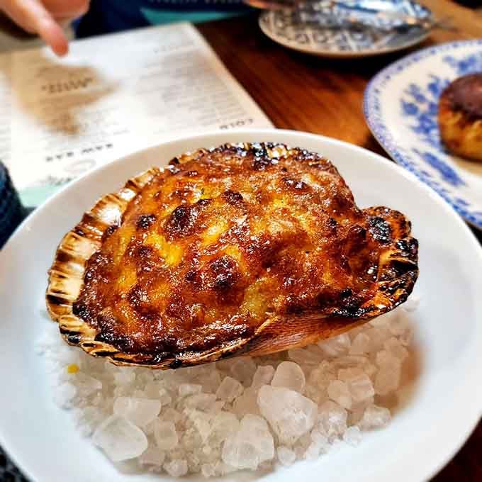 A baked scallop presented on its shell like edible treasure, golden and bubbling with the kind of richness that haunts dreams.
