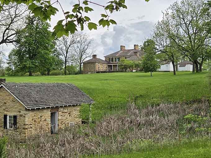 A small stone outbuilding anchors the foreground while the grand mansion watches patiently from the rolling green hillside above.