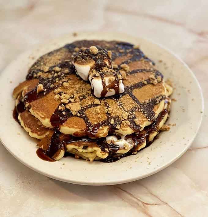 S'mores pancakes drizzled with chocolate and topped with marshmallows. Campfire dreams, diner style, no tent required.