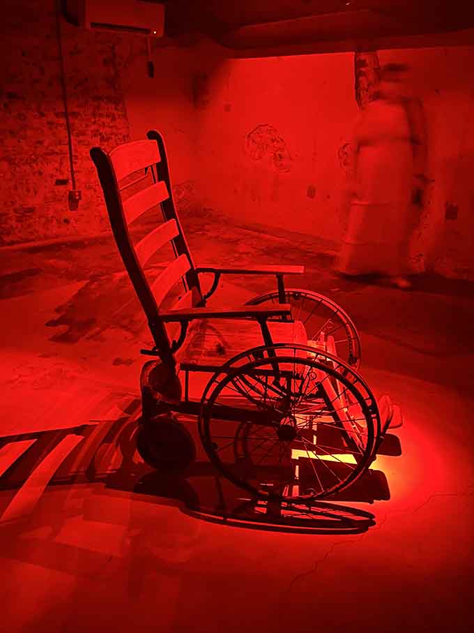 A vintage wheelchair bathed in red light, with ghostly figures sketched across the walls behind it.
