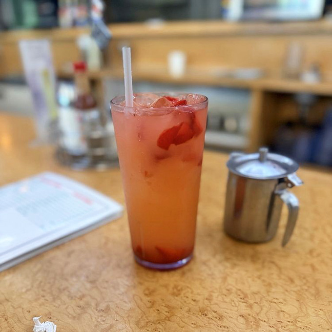 A tall, rosy strawberry lemonade packed with ice and real fruit pieces, sitting pretty at the counter like a summer postcard.