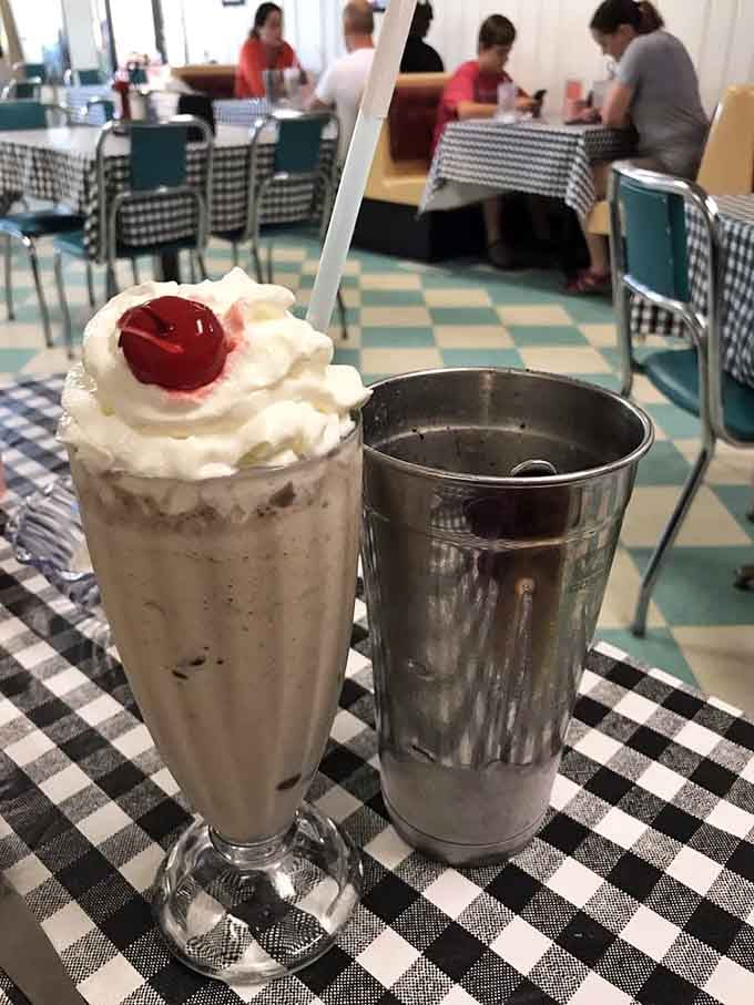 A milkshake served the old-fashioned way, tall glass, metal cup on the side, cherry on top.