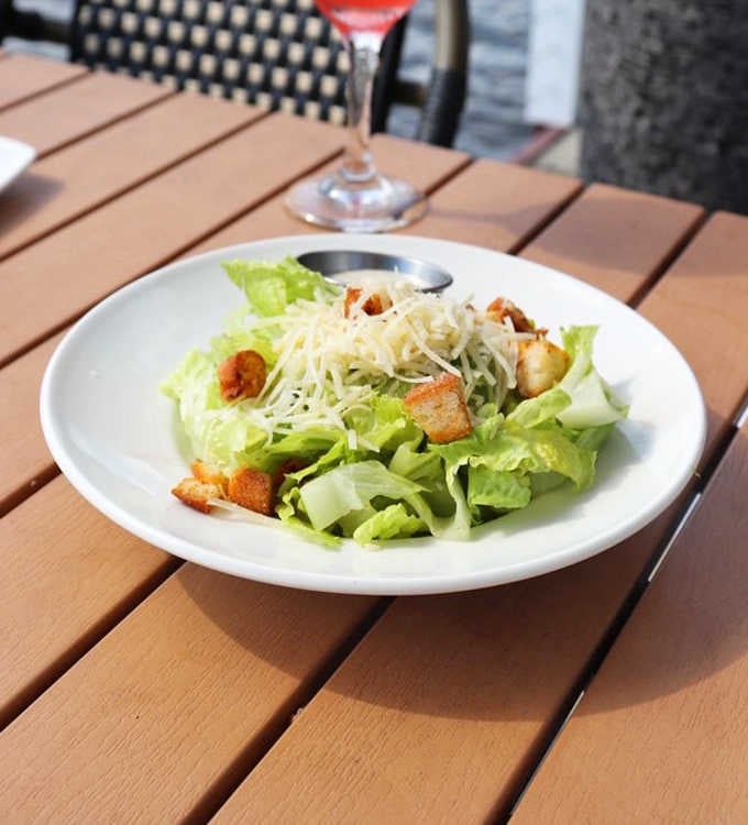 A classic Caesar salad with shredded parmesan and golden croutons, a rosy cocktail glowing softly in the background.