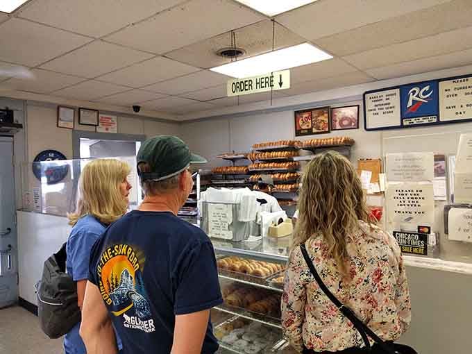 The "Order Here" sign points down, but everyone's eyes are pointed straight at those beautiful donuts on the rack.