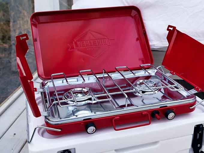 A cherry-red camp stove sitting ready on a cooler. Breakfast in the woods never looked this cheerful or this capable.