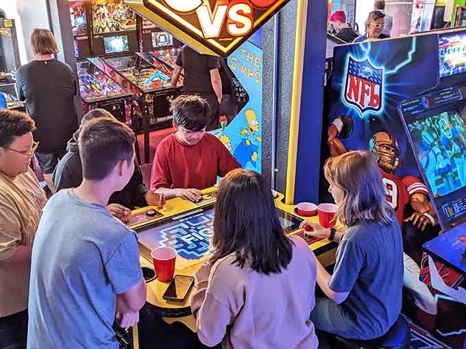 Groups gather around The Simpsons arcade table, red cups in hand, laughter filling the room.