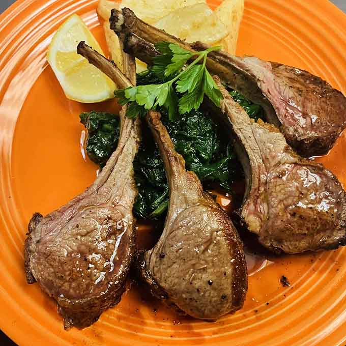 Four perfectly seared lamb chops fanned over wilted spinach on a bold orange plate, dinner just got dramatic.