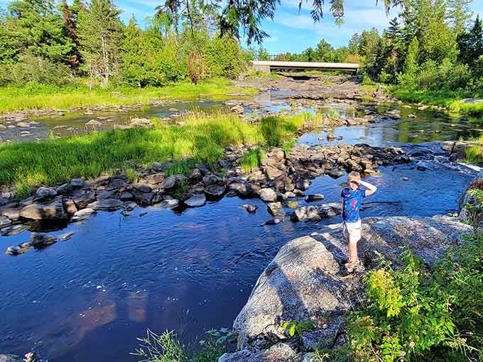 A young explorer takes in the view of the gentle stream and distant bridge during a sunny day of adventure.