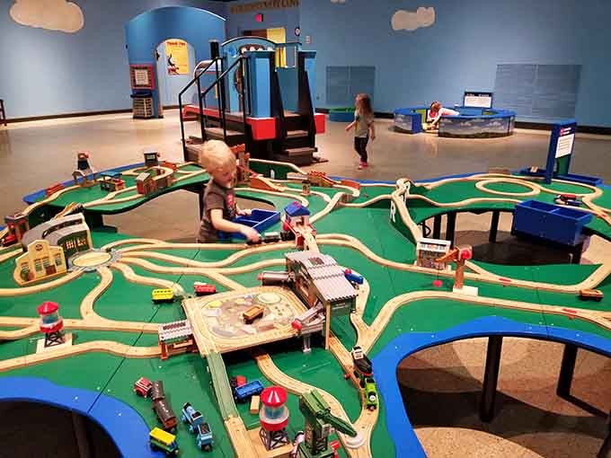 A toddler navigating an enormous wooden train table layout, completely in charge of his own little world of tracks and tiny locomotives.