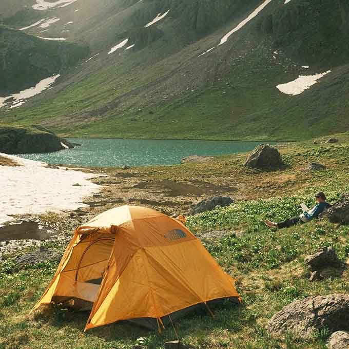An orange tent glows against the green alpine meadow while someone reads beside the lake, living their absolute best life.
