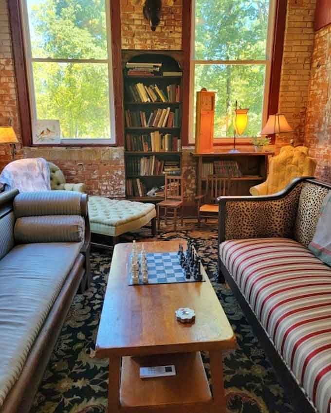 A chess set mid-game on a wooden coffee table, framed by lush green trees and warm brick walls outside tall windows.