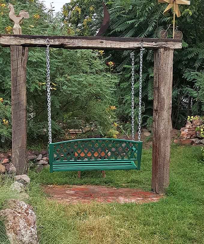 A garden swing hanging from a rustic wooden frame, surrounded by green trees, built specifically for doing absolutely nothing productive.
