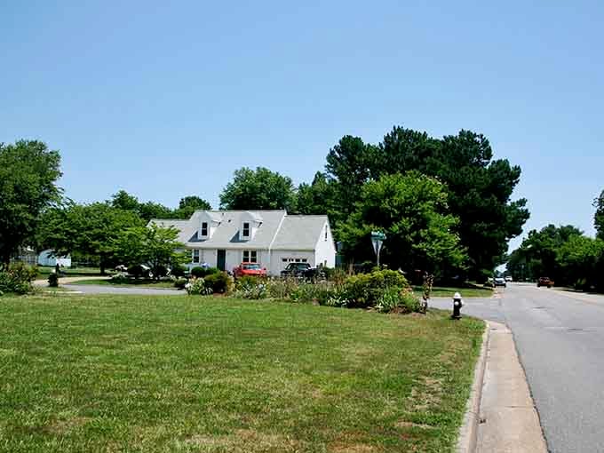 A lovely white suburban home sits peacefully behind a wide, sunlit green lawn on a beautiful, clear blue day.