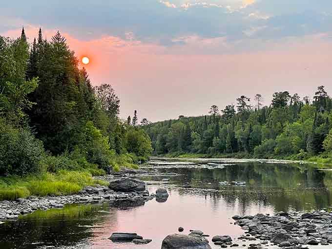 The red sun hangs low in a hazy sky, reflecting softly on the calm surface of the wide, rocky river.