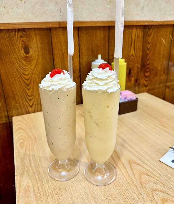 Two tall milkshakes topped with whipped cream and cherries, because every great deli meal deserves a proper, indulgent finale.