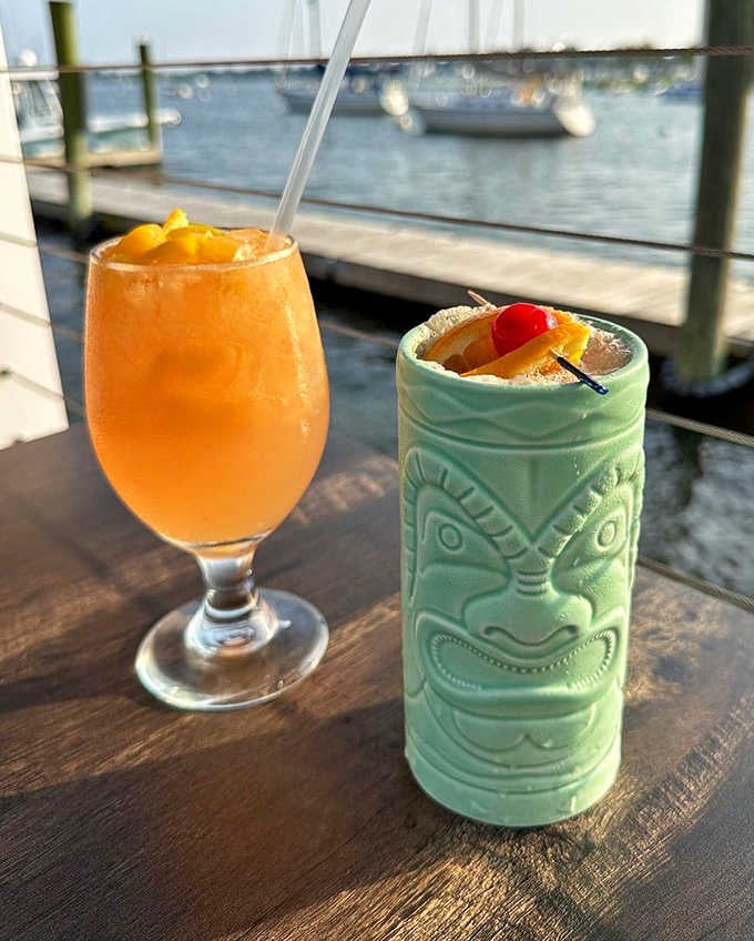 A fruity cocktail in a stemmed glass beside a tiki mug, sailboats drifting lazily across the harbor behind them.