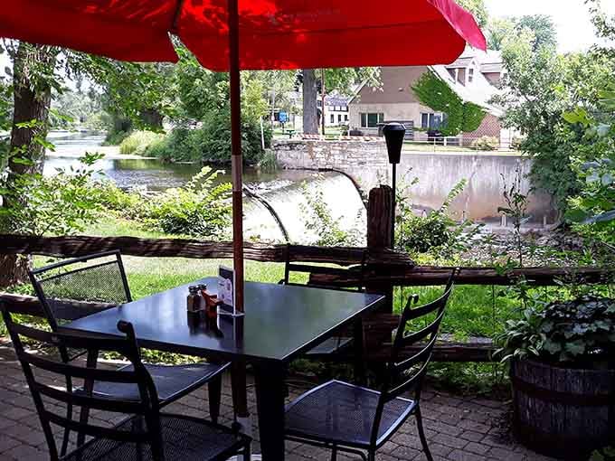 A patio table with a red umbrella, Cedar Creek rolling past in the background &mdash; lunch with a waterfall view, no passport required.