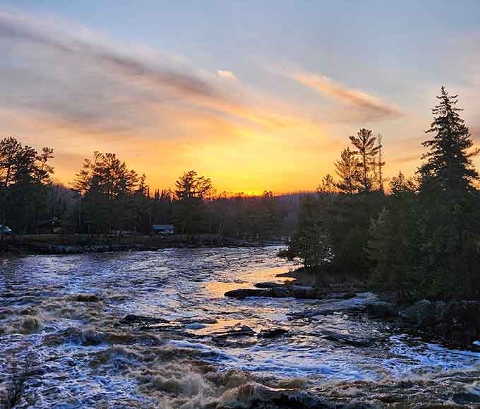Vibrant orange and yellow hues fill the horizon as twilight falls over the rushing rapids and the quiet northern woods.