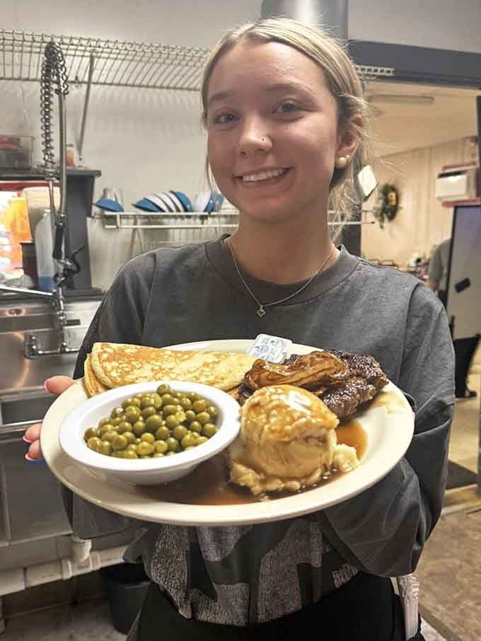 A server proudly presenting a loaded plate of pancakes, peas, mashed potatoes, and gravy straight from the kitchen &mdash; that smile says it all.