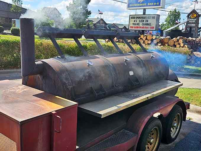 Blue smoke curling off a massive trailer smoker with a sign reading "Voted Springfield's Best BBQ" visible right behind it. Case closed.