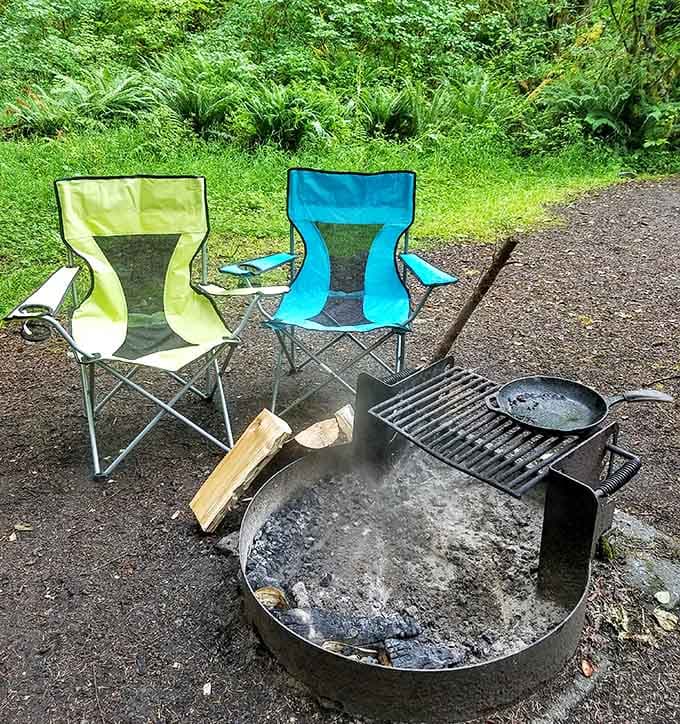 A cast iron skillet, a smoldering fire ring, and two empty chairs &mdash; dinner is almost ready, friend.