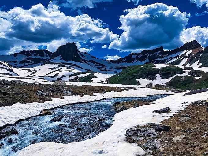 A rushing snowmelt stream cuts through a white winter landscape beneath dramatic clouds, raw and alive in the most elemental way possible.