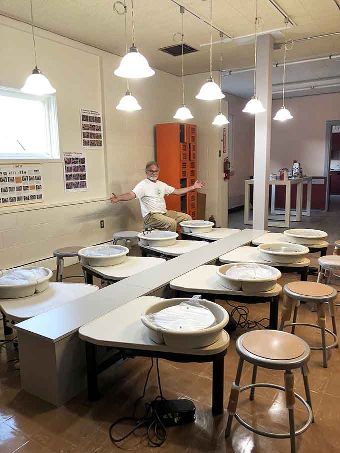 A cheerful instructor spreads his arms wide among rows of pottery wheels, looking like a man who has genuinely found his happy place in clay.