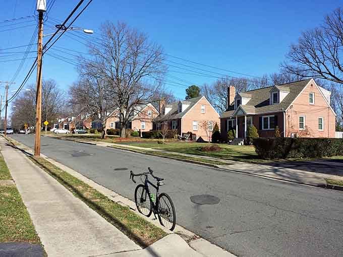 A lone bicycle rests along the sidewalk of a peaceful, tree-lined residential street filled with classic brick houses.
