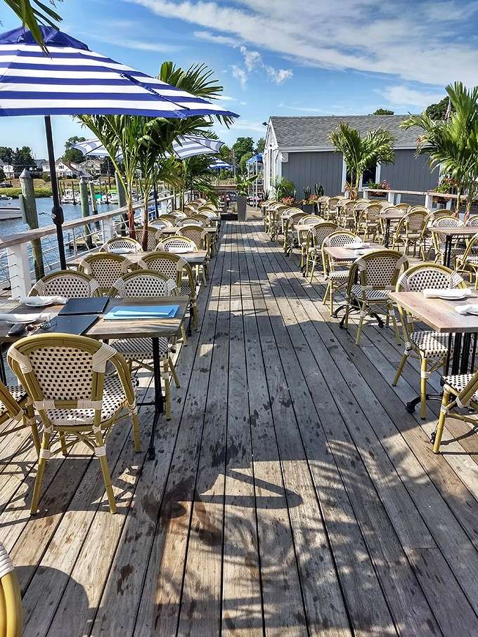 A sun-drenched deck lined with bistro chairs, striped umbrellas, and palm trees swaying beside the calm Norwalk harbor.