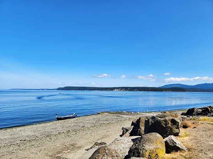 One lone rowboat rests on a sun-drenched rocky shore, with glassy blue water stretching toward distant hills under a sky that's showing off.