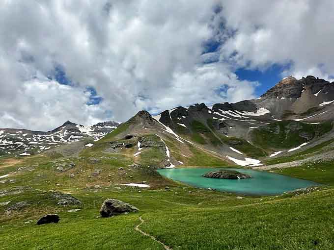 Dramatic clouds roll over snow-dusted peaks while that jewel-toned lake glows quietly below, completely unbothered by the drama above.