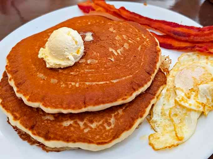 Three golden pancakes stacked tall, crowned with butter, flanked by crispy bacon and sunny eggs. Morning perfection, full stop.