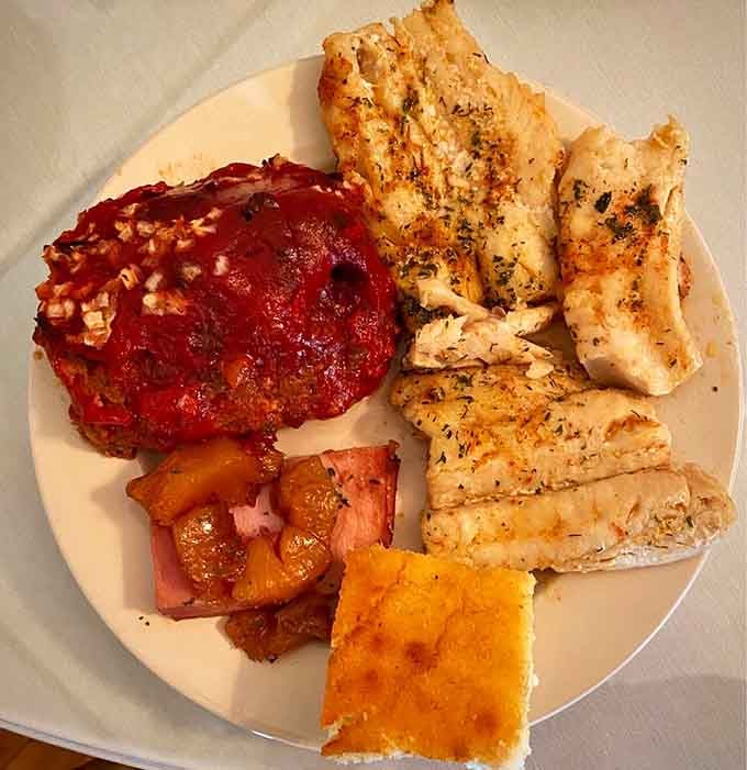 Herb-seasoned baked fish alongside glazed meatloaf and sweet cooked fruit, comfort food that means serious business without showing off.