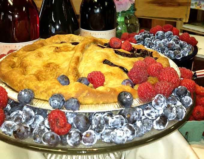 A berry pie surrounded by fresh raspberries and blueberries, with farm wine bottles standing nearby like very proud parents.