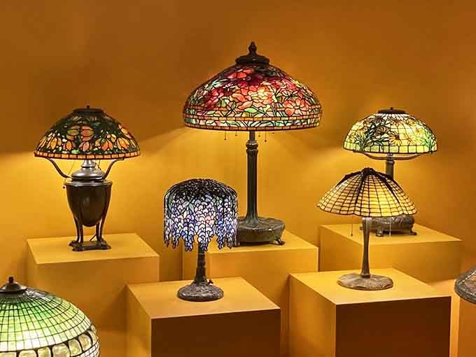 Five Tiffany lamps glowing against a golden wall, each one a tiny stained-glass universe you'd absolutely rearrange your living room to accommodate.