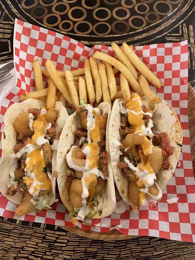 Three shrimp tacos loaded with cheese sauce and fresh toppings, sitting on red checkered paper like edible confetti.