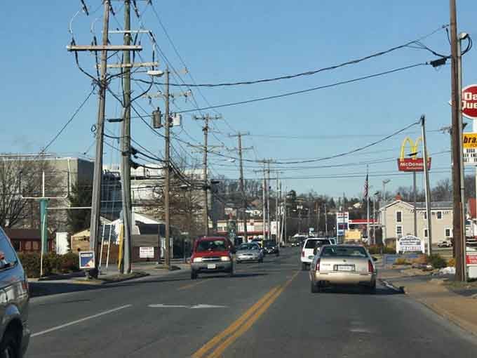 Drive down this busy town road lined with local shops, familiar signs, and utility poles under a clear blue sky.